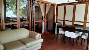 Quarto com sofá, mesa de jantar e cadeiras, e vista para o mar através de grandes janelas.