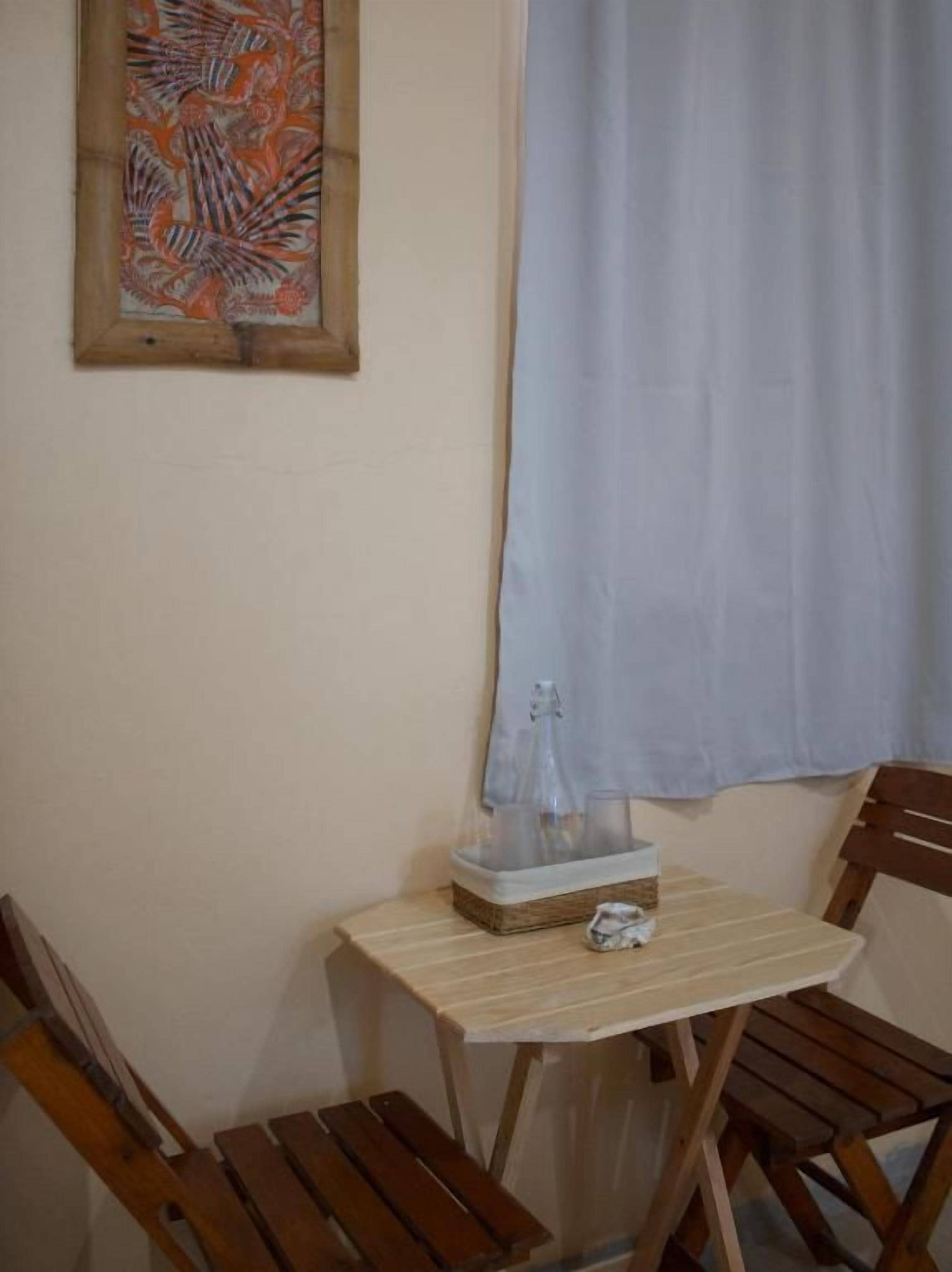 Quarto pequeno com mesa de madeira, duas cadeiras, uma obra de arte emoldurada na parede e uma janela com cortina branca.