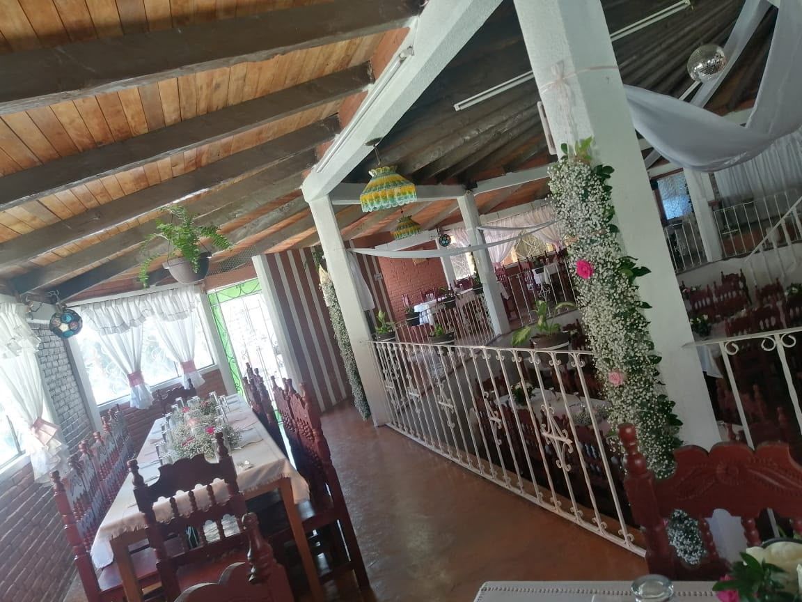 Espaço de refeições interno com vigas de madeira, colunas brancas e uma grade com plantas pendentes. Mesas dispostas com toalhas brancas e arranjos florais.