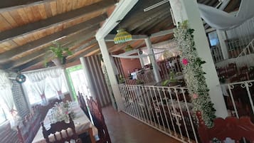 Espaço de refeições interno com vigas de madeira, colunas brancas e uma grade com plantas pendentes. Mesas dispostas com toalhas brancas e arranjos florais.
