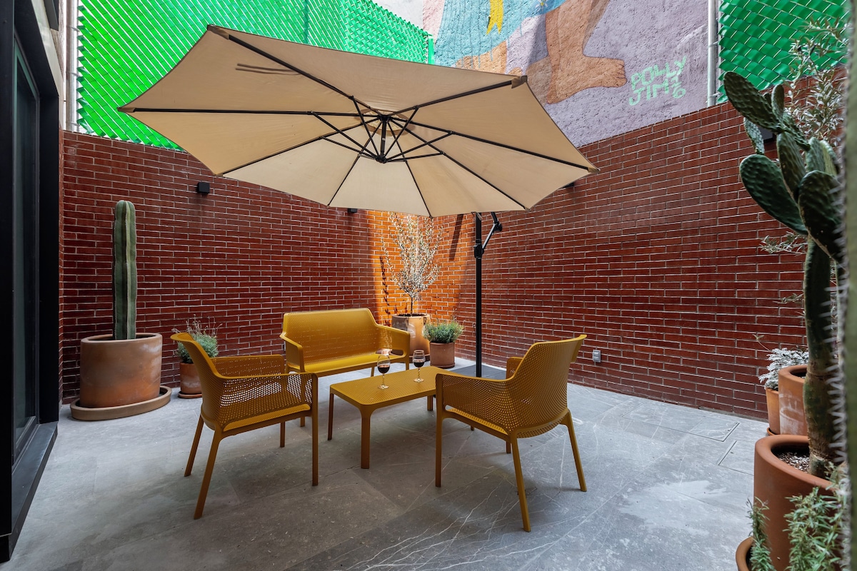 Área de pátio com muro de tijolos, mesa e cadeiras trançadas amarelas, um guarda-sol bege e plantas em vasos.