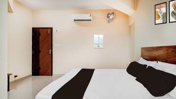 Quarto de hotel com uma cama, porta de madeira, aparelho de ar condicionado, pequena janela e ventoinha fixada na parede.