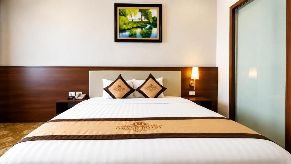 Quarto de hotel com uma cama grande, cabeceira com logotipo, mesa de cabeceira com abajur e um quadro na parede.