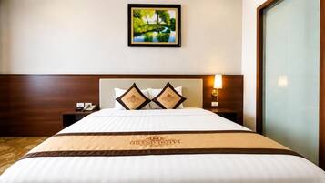 Quarto de hotel com uma cama grande, cabeceira com logotipo, mesa de cabeceira com abajur e um quadro na parede.