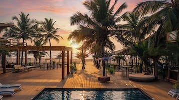 Área de piscina com espreguiçadeiras e deck de madeira, com vista para uma praia com palmeiras.