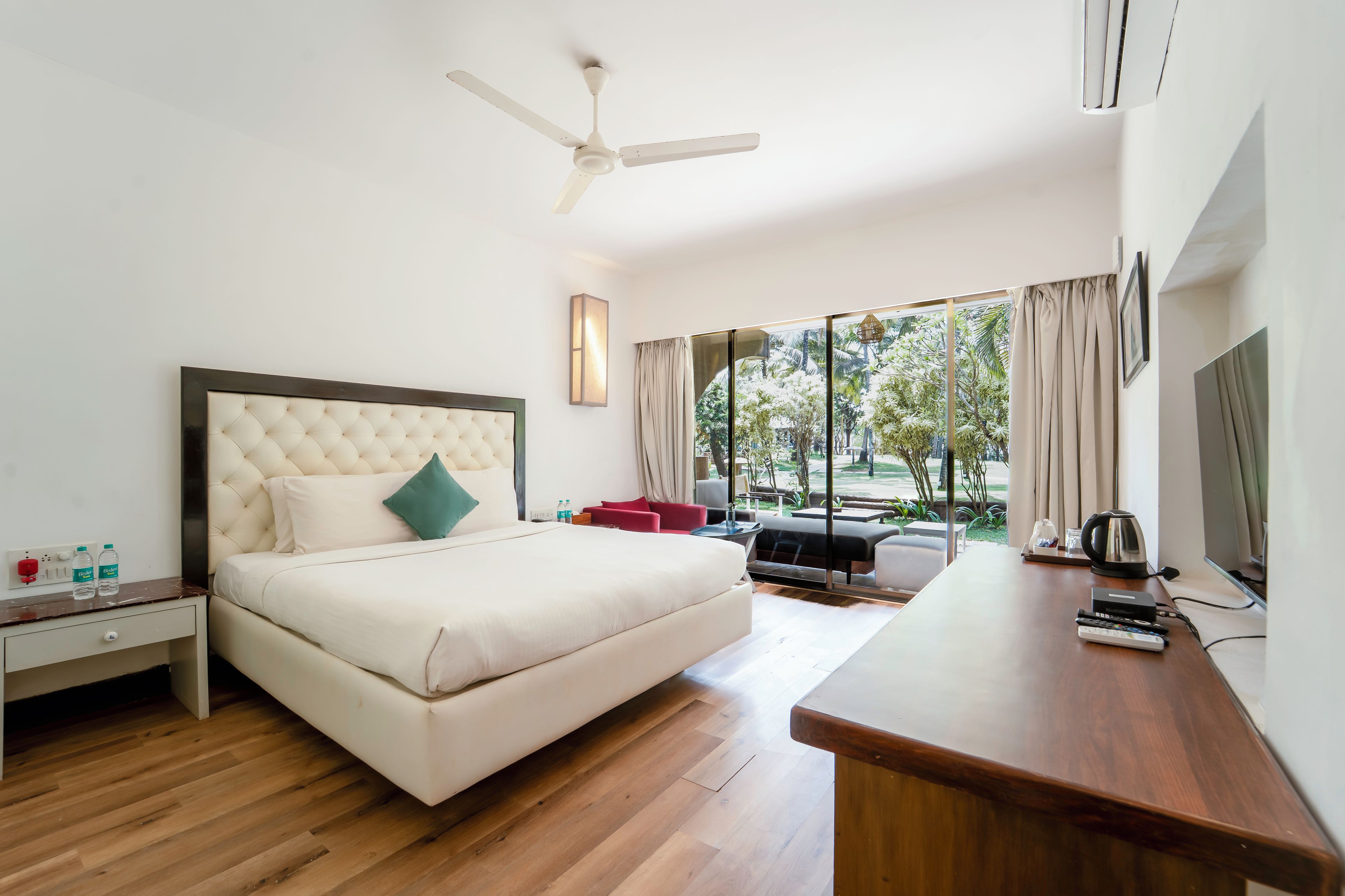 Quarto de hotel com cama, mesa de cabeceira, escrivaninha, cadeira, televisão e ventoinha no teto.