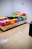 Cama com colcha floral e várias almofadas, piso de azulejos e parede lisa com tomada.