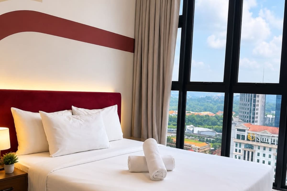 Quarto de hotel com uma cama grande, mesa de cabeceira, abajur e vista da cidade por grandes janelas.