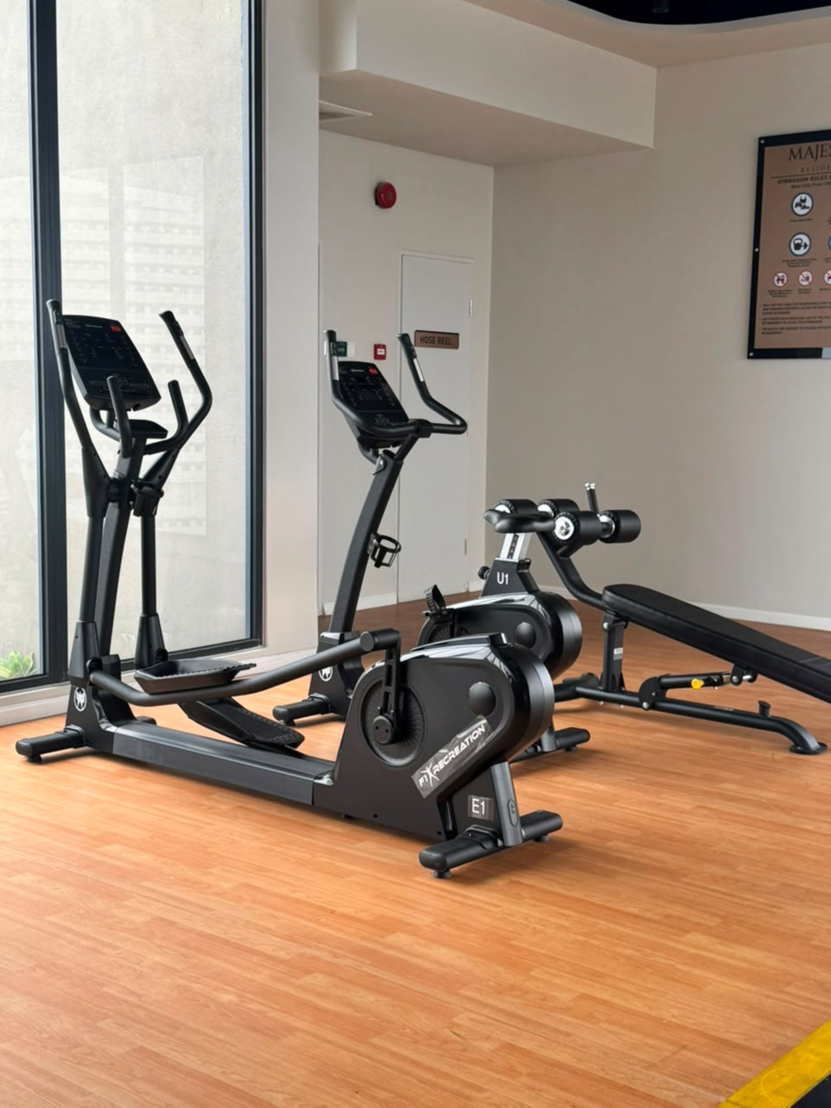 Academia com equipamentos de ginástica, incluindo bicicleta ergométrica e elíptico, em um espaço amplo com janelas grandes e piso de madeira.