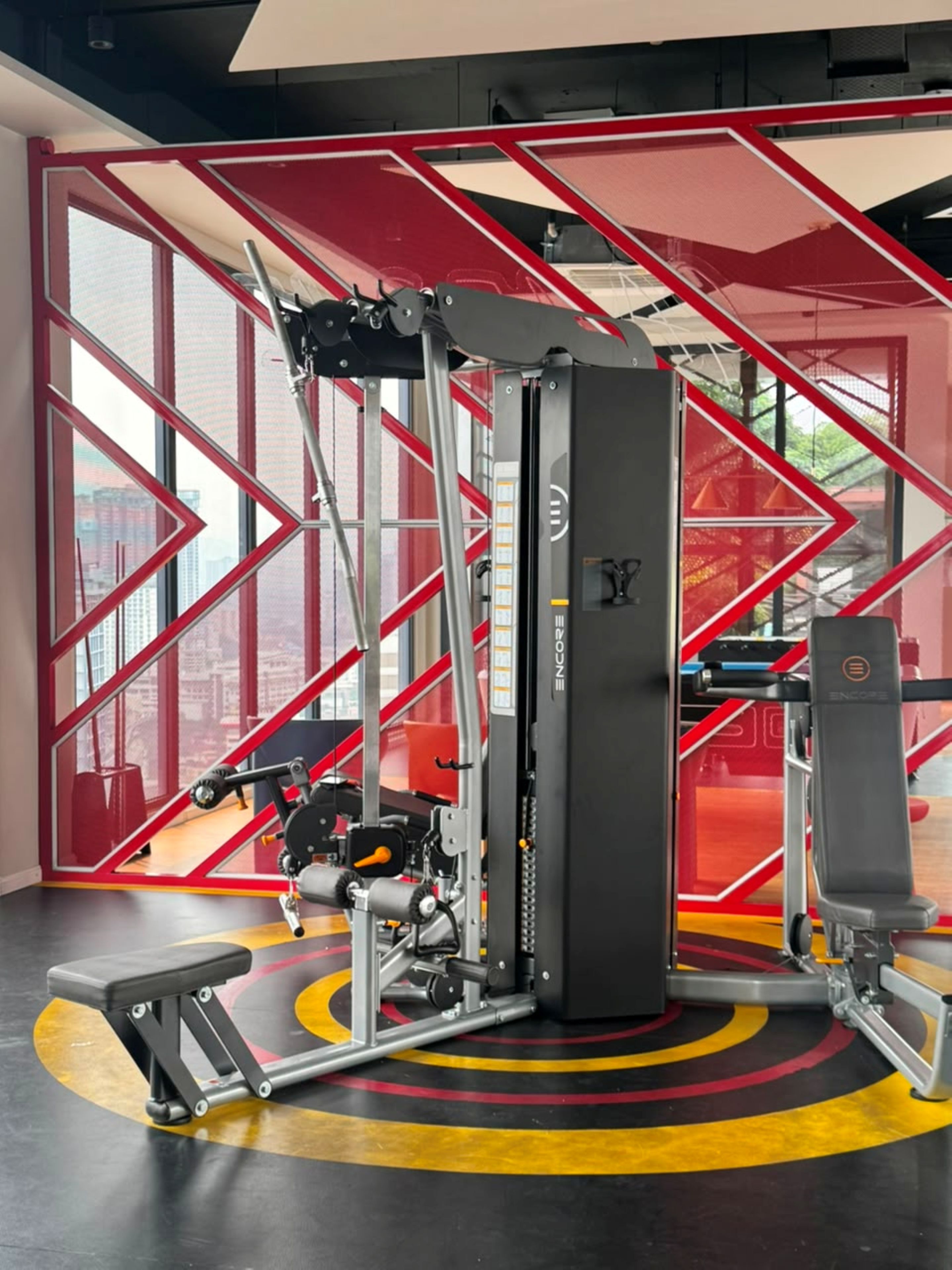 Uma academia com suporte para agachamento, banco de musculação e design geométrico preto e vermelho na parede.