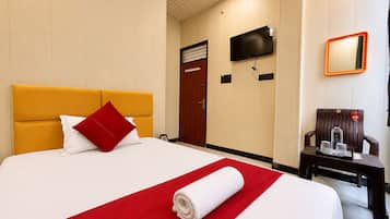Quarto de hotel com cama, edredom vermelho e branco, toalha branca, mesa de cabeceira de madeira, espelho e televisão na parede.