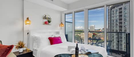 Quarto moderno com cama grande, mesa de cabeceira, uma cadeira e varanda com vista para a cidade.
