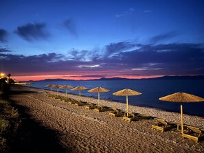 Uma praia com guarda-sóis e espreguiçadeiras, um litoral rochoso e um pôr do sol sobre a água.