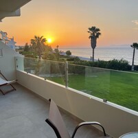 Varanda com vista para o pôr do sol sobre o mar, área gramada e uma palmeira.