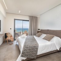 Quarto de hotel com uma cama grande, área de estar com mesa e cadeiras, e vista para o mar através de uma porta de correr.