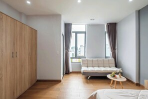 Quarto moderno com guarda-roupa espaçoso, sofá-cama, mesa redonda pequena com uma tigela de frutas e vista para a cidade através de uma janela grande.