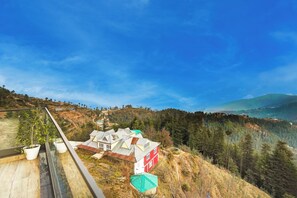 Paisagem montanhosa com um edifício vermelho e branco, possivelmente uma hospedaria, rodeada por árvores e um céu claro.