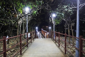 Uma passarela noturna com grades de metal e iluminação suspensa, conduzindo a uma construção ao longe.