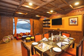 Uma área de refeições com teto de madeira, uma janela grande com cortinas, uma televisão na parede e uma mesa posta para refeição.