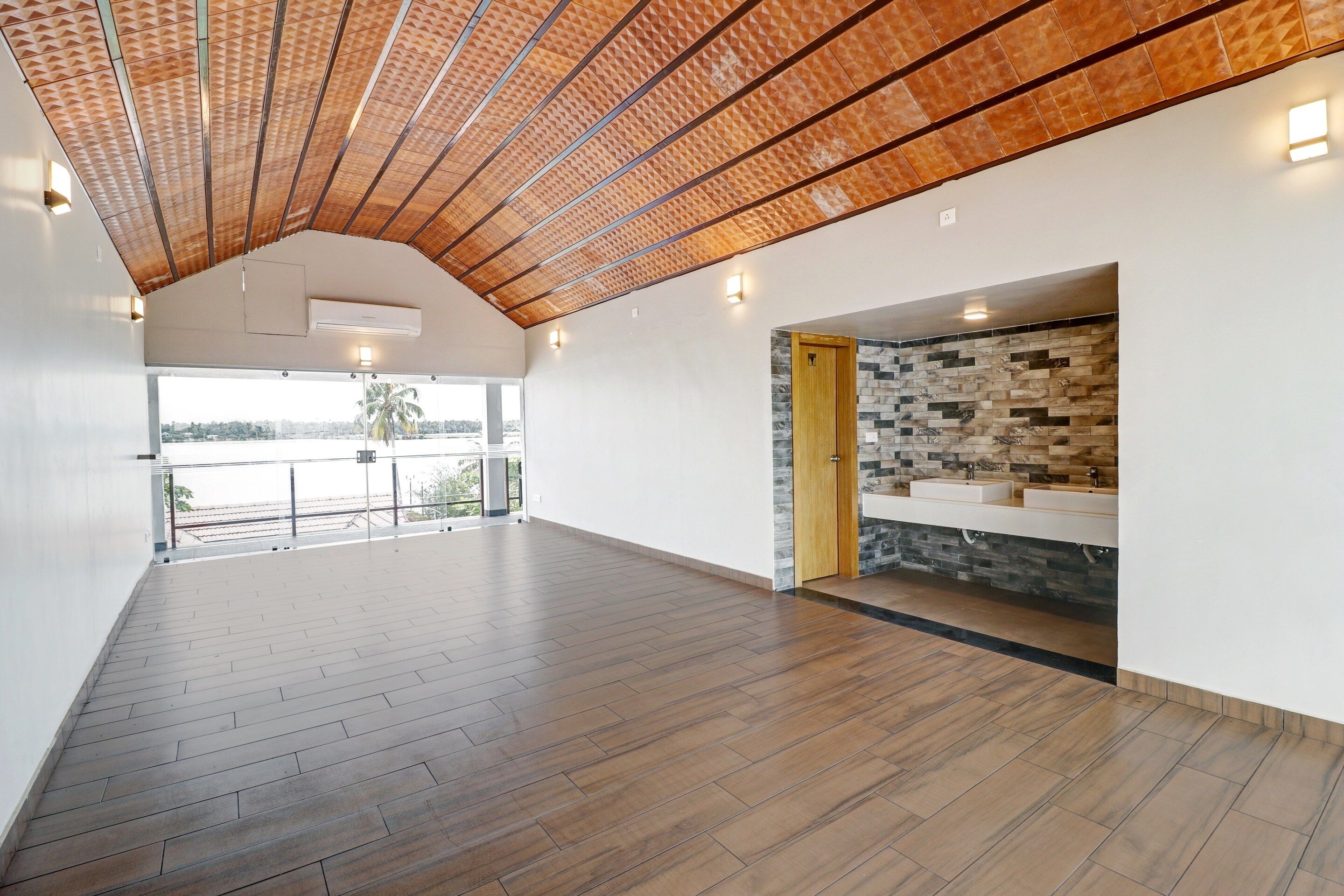 Quarto espaçoso com piso de madeira, teto inclinado com padrão geométrico e vista para palmeiras através de grandes janelas.