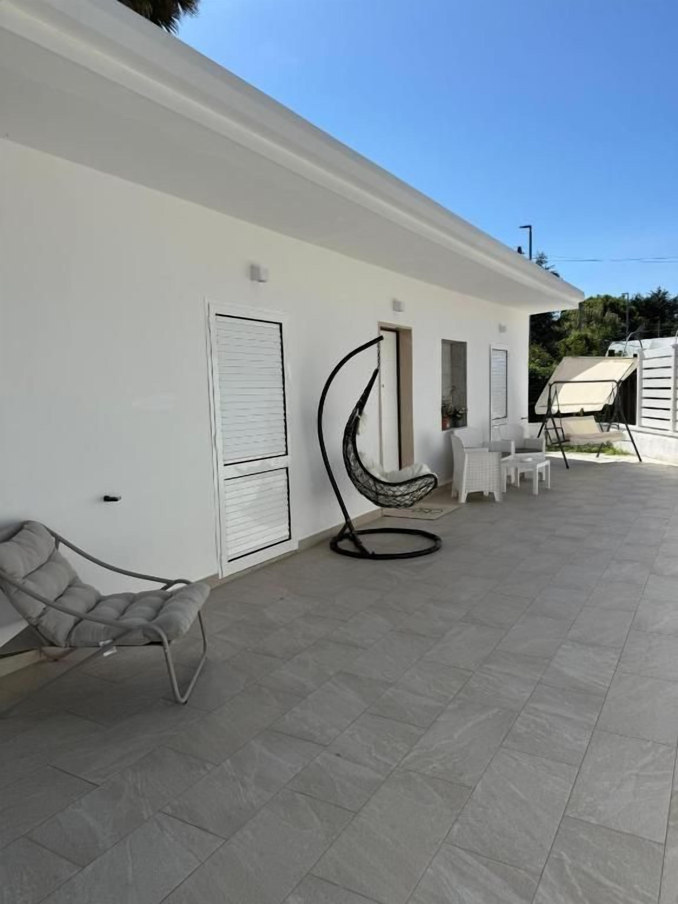 Uma área de estar ao ar livre com um edifício branco, uma cadeira suspensa, uma poltrona estofada e uma pequena mesa.