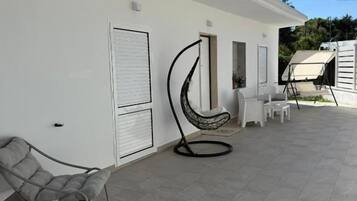 Uma área de estar ao ar livre com um edifício branco, uma cadeira suspensa, uma poltrona estofada e uma pequena mesa.