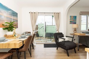 Uma área de refeições moderna com uma mesa de madeira posta para quatro pessoas, uma cadeira preta e vista para um espaço ao ar livre através de uma porta de vidro.
