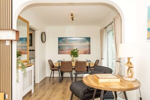 Uma sala de jantar com mesa de madeira, cadeiras e um quadro de um pôr do sol.