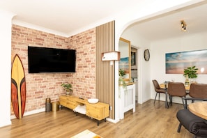 Uma sala de estar moderna com parede de tijolos, uma TV de tela plana, um console de madeira, uma área de jantar com mesa redonda e um quadro grande.