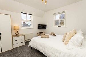 Quarto com cama, televisão, escrivaninha com luminária e duas janelas.