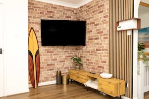Sala de estar com parede de tijolos, televisão montada, uma prancha de surf, armário de madeira e uma planta.