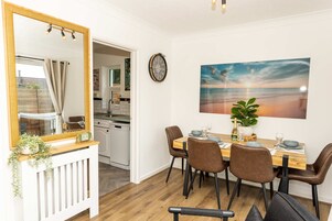 Uma área de refeições com mesa de madeira, cadeiras marrons, uma pintura grande de um pôr do sol e uma cozinha ao fundo.