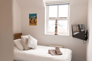 Quarto com cama, almofadas, TV na parede, um quadro e janela com vista para um farol.