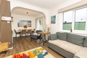 Uma sala de estar moderna com um sofá cinzento, um espaço de refeições com cadeiras, um tapete colorido e obras de arte nas paredes.