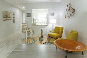 Uma sala de estar com sofá, poltrona e uma cadeira laranja singular. Há um tapete estampado, uma mesinha com uma vela e um abajur.