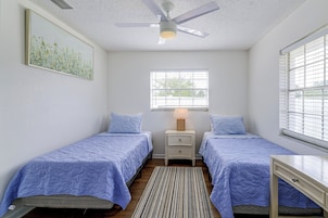 Quarto com duas camas de solteiro, ventoinha no teto, janela com persianas e mesa de cabeceira com abajur.