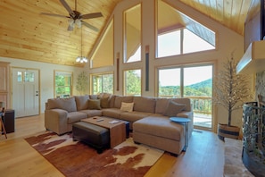 Uma sala de estar ampla com um sofá seccional grande, uma mesa de centro e uma lareira. Há várias janelas que proporcionam luz natural e vistas para o exterior.