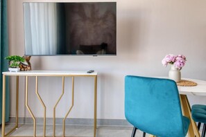 Uma sala de estar moderna com uma TV de tela plana na parede, uma mesa branca com moldura dourada, uma cadeira azul e um vaso com flores cor-de-rosa.