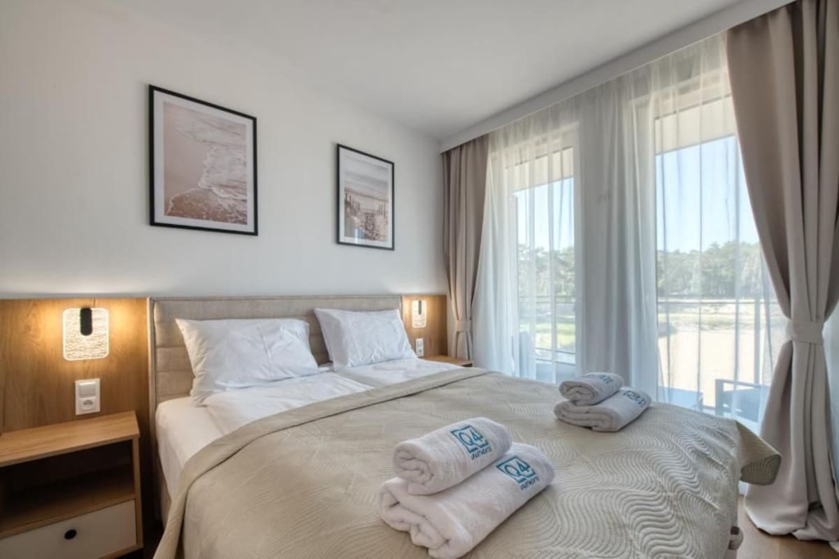 Quarto de hotel com uma cama grande, duas almofadas, mesa de cabeceira e uma janela com cortinas.