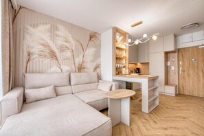 Uma sala de estar moderna com um sofá seccional, uma pequena mesa redonda e uma cozinha planejada com armários de madeira e uma luminária pendente.