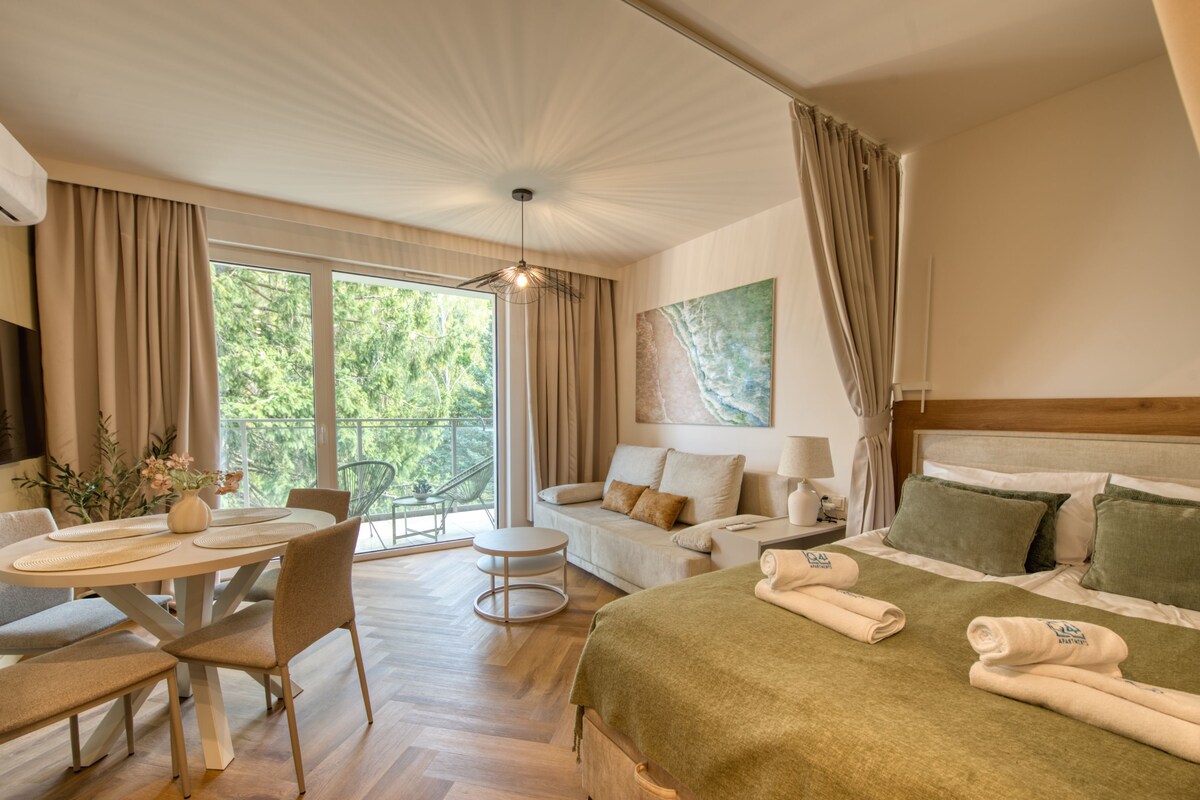 Quarto de hotel com varanda, cama com almofadas verdes, mesa redonda com cadeiras e um quadro na parede.