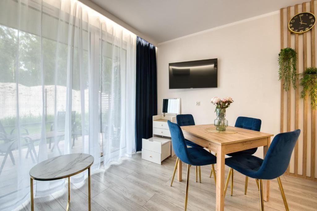 Uma sala de jantar moderna com mesa de madeira, cadeiras azuis, televisão de tela plana e janela com cortinas leves.