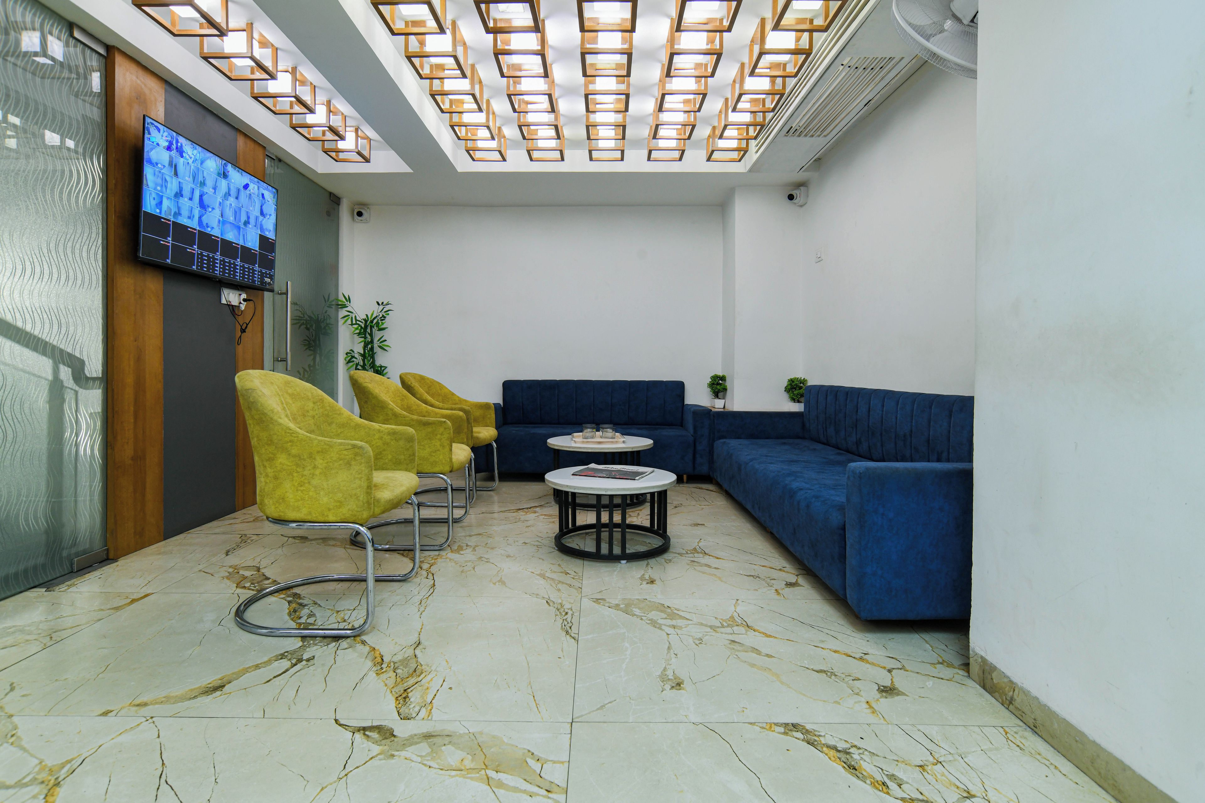 Uma área de espera moderna com piso de mármore, cadeiras amarelas, um sofá azul e uma TV de tela plana fixada na parede.