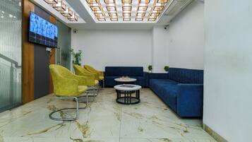 Uma área de espera moderna com piso de mármore, cadeiras amarelas, um sofá azul e uma TV de tela plana fixada na parede.