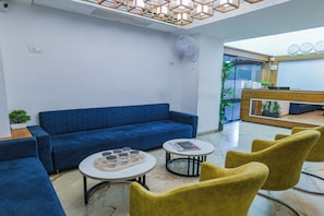 Uma área de estar moderna com um sofá azul, cadeiras amarelas, uma mesa redonda pequena com copos e um ventilador na parede.