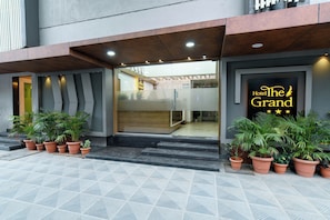 A entrada do Hotel The Grand com design moderno, apresentando uma porta de vidro, plantas em vasos e uma placa com o nome do hotel.