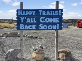 Placa com o texto 'HAPPY TRAILS! Y'ALL COME BACK SOON!' em ambiente rural, com chão de cascalho e céu claro.