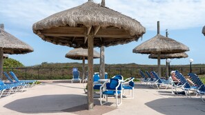 Uma área de estar à beira-mar, com guarda-sóis de palha e espreguiçadeiras azuis.