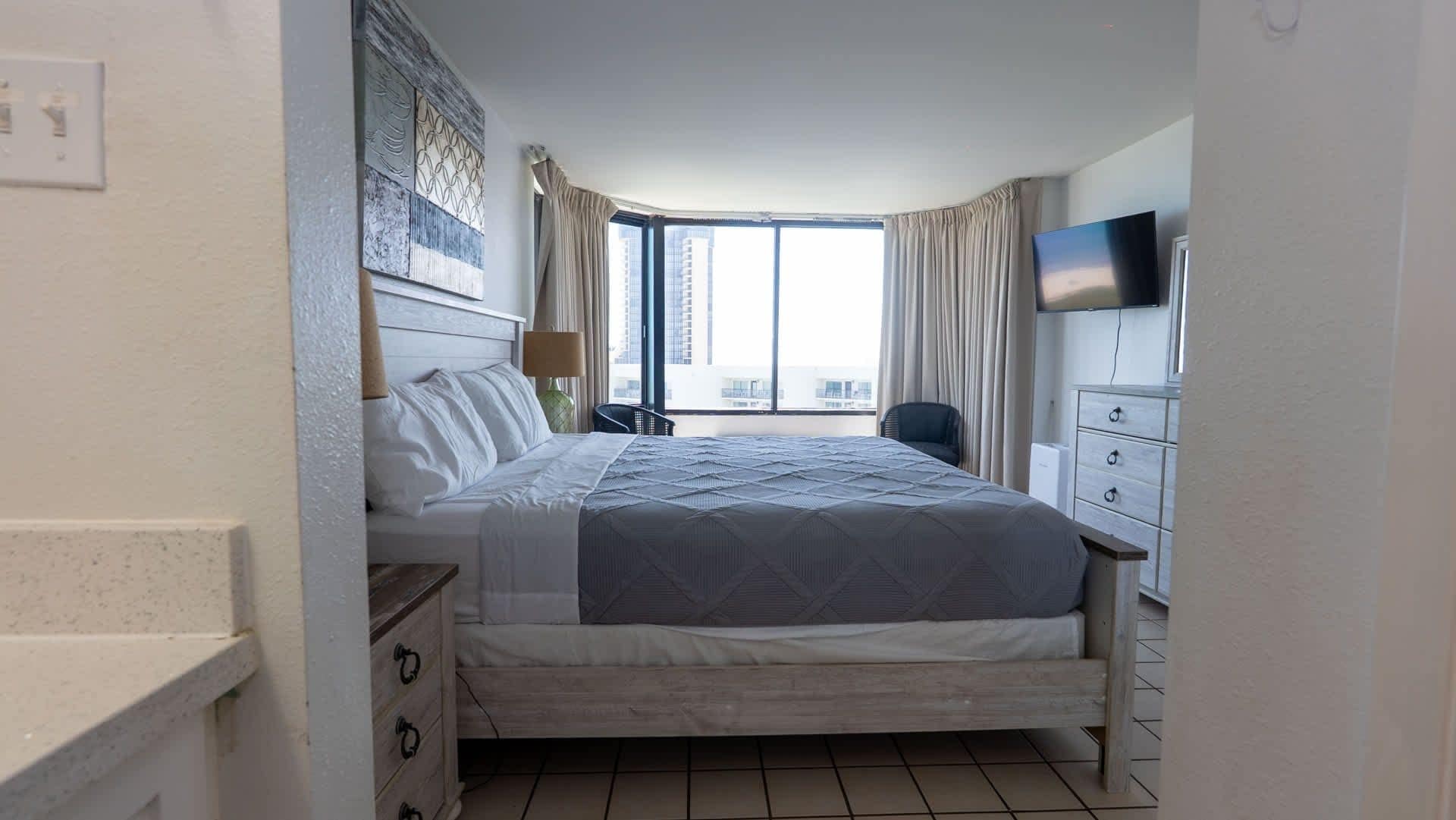 Quarto de hotel com cama, televisão, escrivaninha e janela com cortinas.