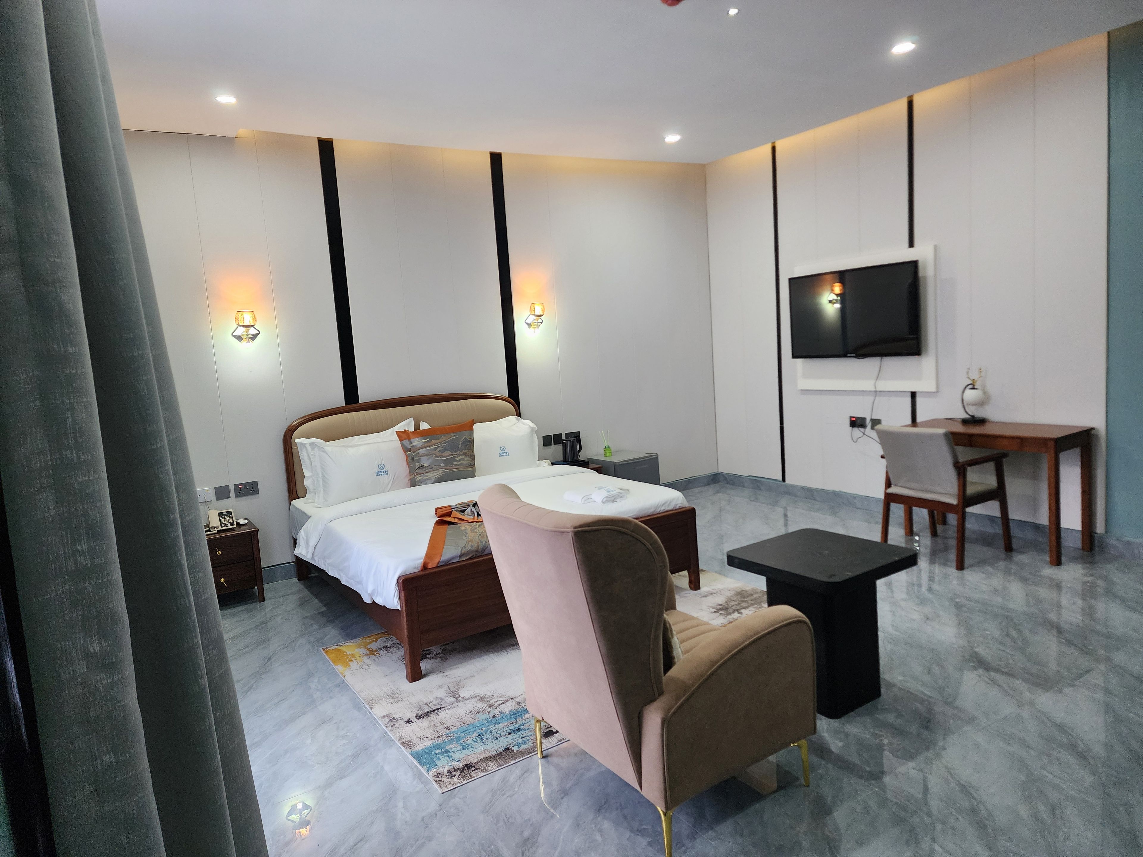 Quarto de hotel com cama, mesa de cabeceira, poltrona, escrivaninha, televisão e pequena mesa com abajur.